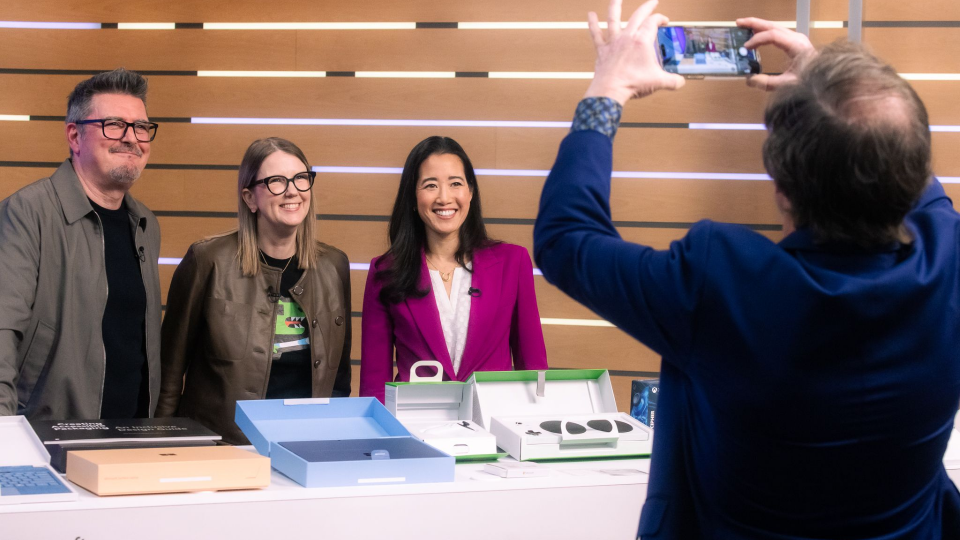 Four people are in the image. Three of them are standing behind a table, and one person is taking a photo of them. The person taking the photo is wearing a blue suit and holding a smartphone. The three people behind the table are smiling. The man on the left is wearing glasses and a brown jacket over a black shirt. The woman in the middle is wearing glasses and a brown jacket over a black shirt with a green design. The woman on the right is wearing a magenta blazer over a white top. The table has several boxes on it, and the word "Microsoft" is visible on the side. The background consists of horizontal wooden panels.